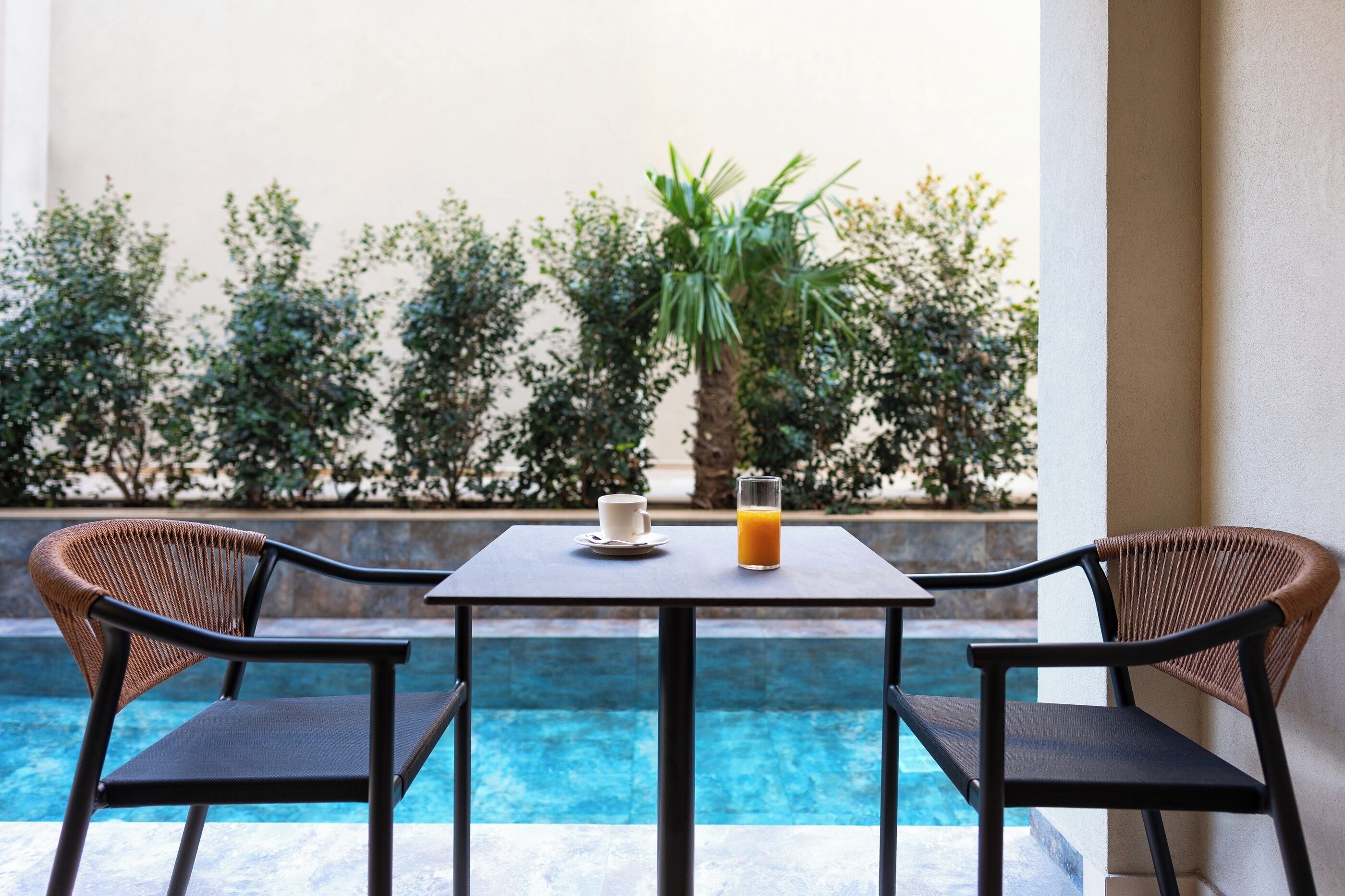 Mesa com duas cadeiras de vime, um copo de suco de laranja e uma xícara branca, com vista para uma piscina e vegetação.
