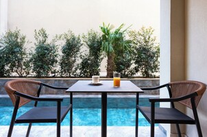 Mesa com duas cadeiras de vime, um copo de suco de laranja e uma xícara branca, com vista para uma piscina e vegetação.