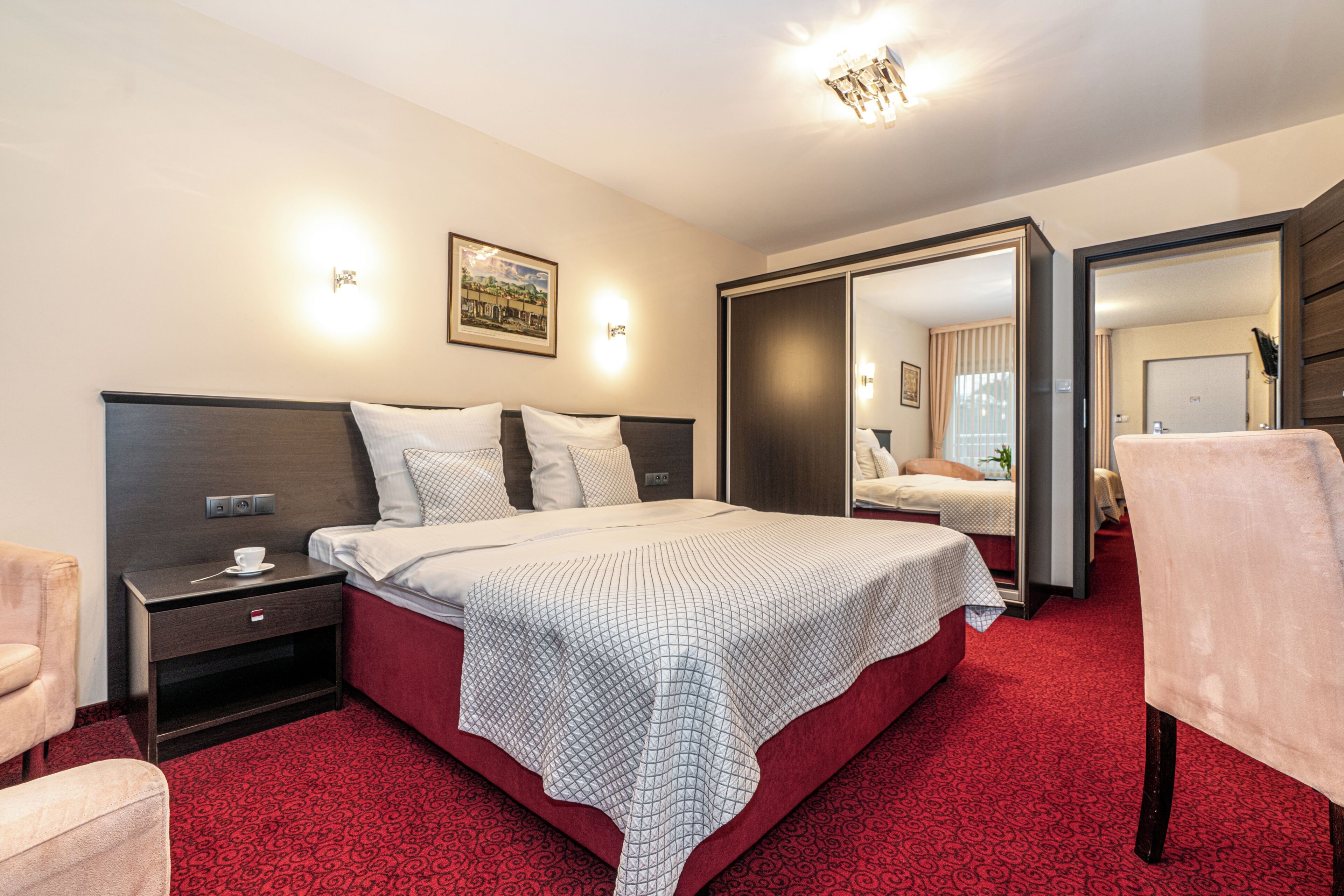 Quarto de hotel com uma cama grande, uma mesinha de cabeceira, uma cadeira, um espelho e um tapete vermelho.