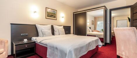 Quarto de hotel com uma cama grande, uma mesinha de cabeceira, uma cadeira, um espelho e um tapete vermelho.