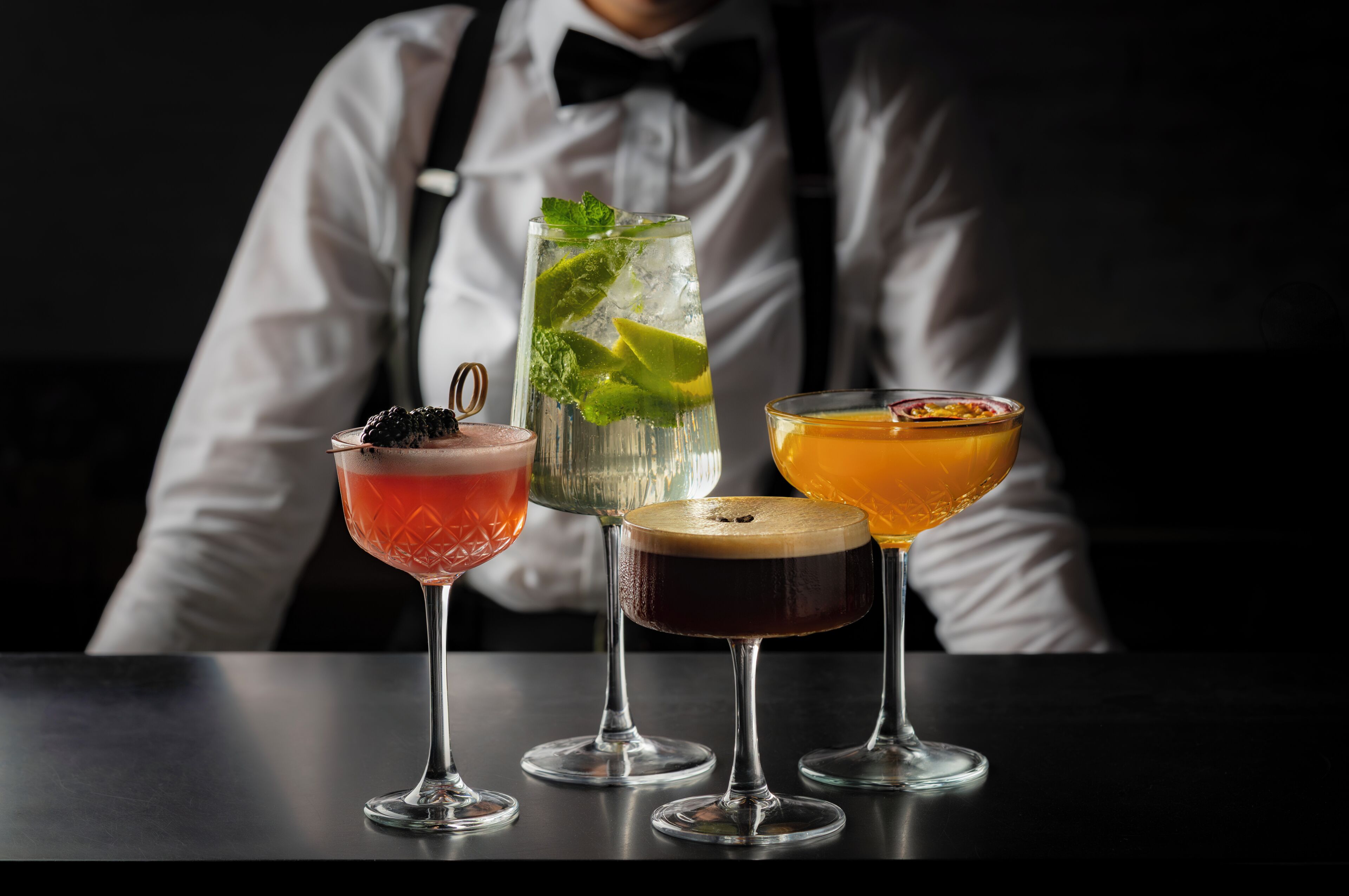 Quatro coquetéis em um balcão de bar, com uma pessoa ao fundo.