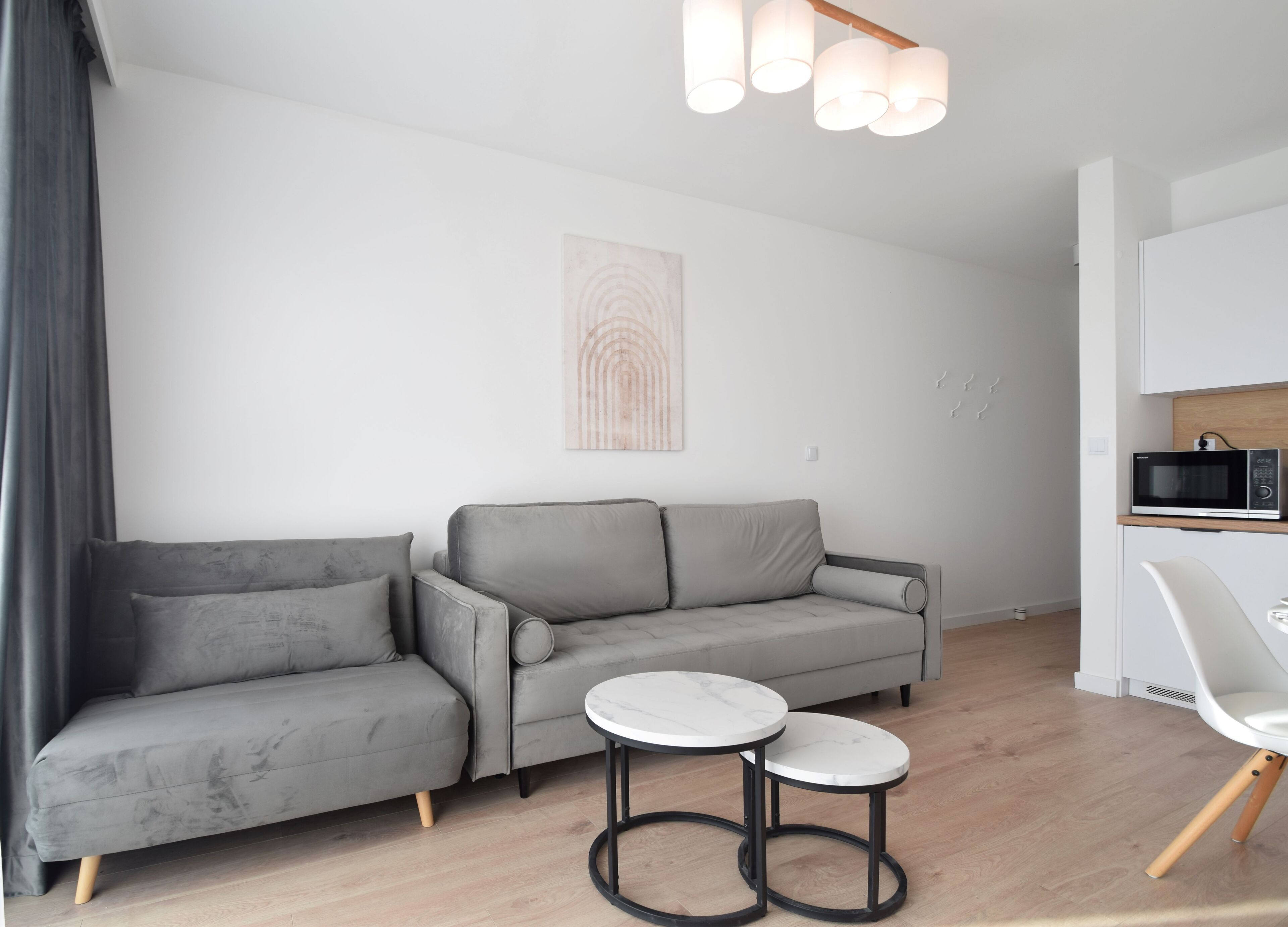 Uma sala de estar moderna com um sofá cinzento, duas mesas redondas, soalho de madeira e uma área de cozinha com micro-ondas.