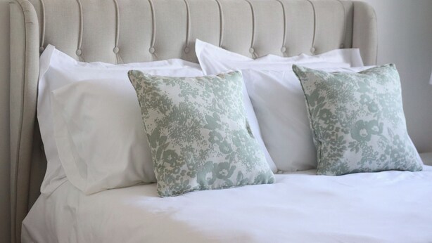 Cama com cabeceira estofada, roupa de cama branca e duas almofadas estampadas.