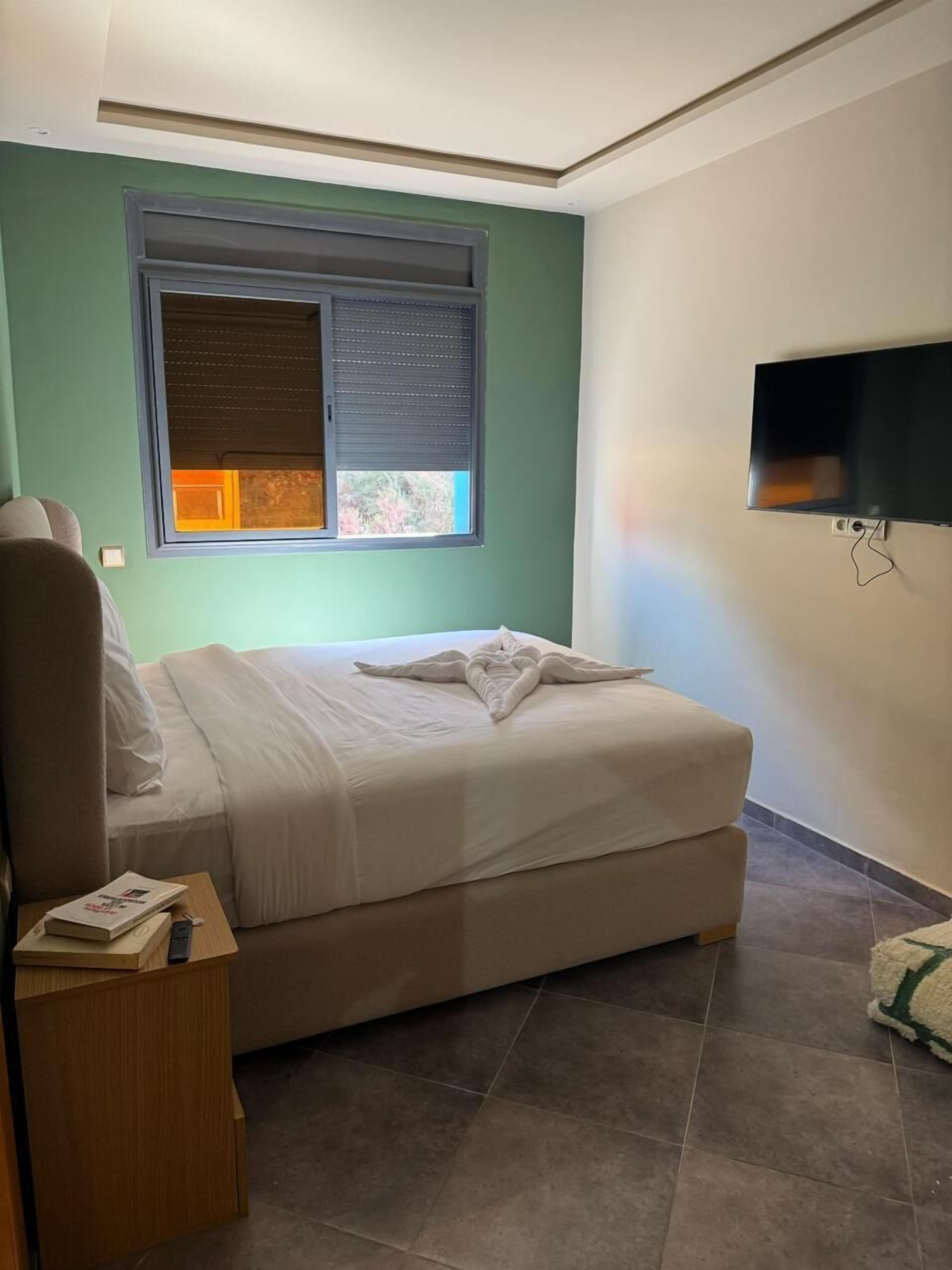 Quarto com cama, janela com persianas, televisão de tela plana na parede e mesa de cabeceira com um livro.