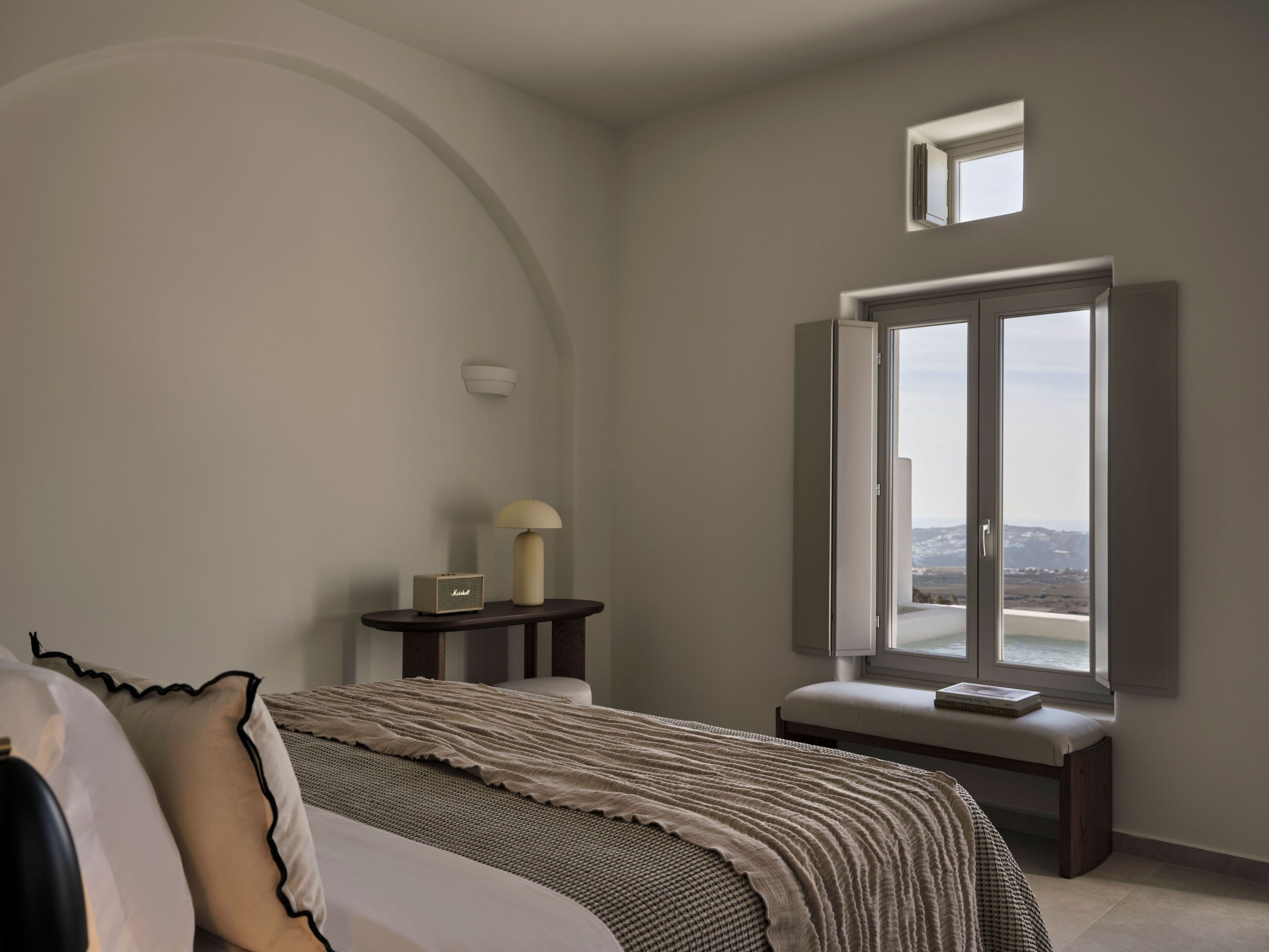 Quarto com uma cama grande, mesa de cabeceira com luminária, mesinha com rádio e janela com vista para o exterior.