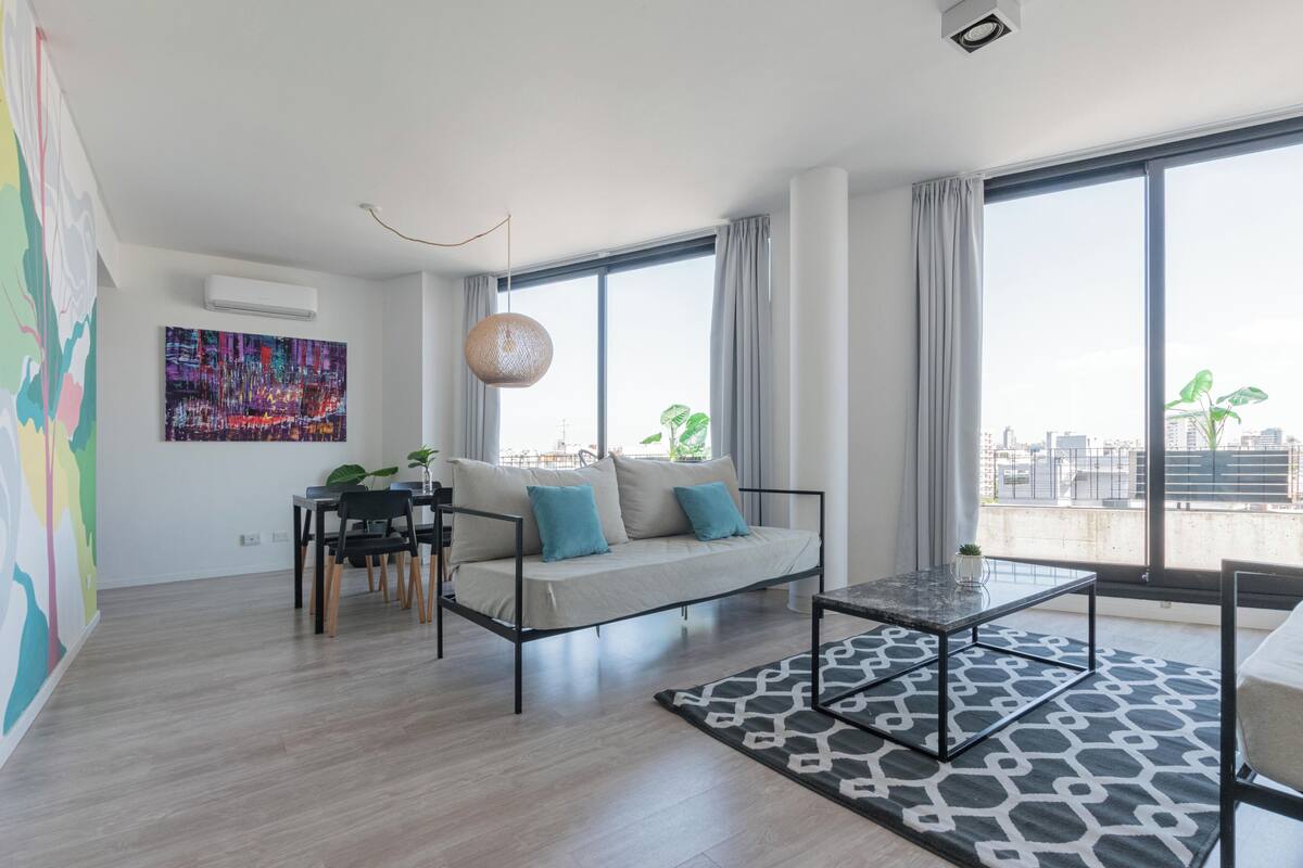 Uma sala de estar moderna com sofá, mesa de centro e mesa de jantar. Há uma janela grande com cortinas, um lustre e um quadro abstrato colorido na parede.