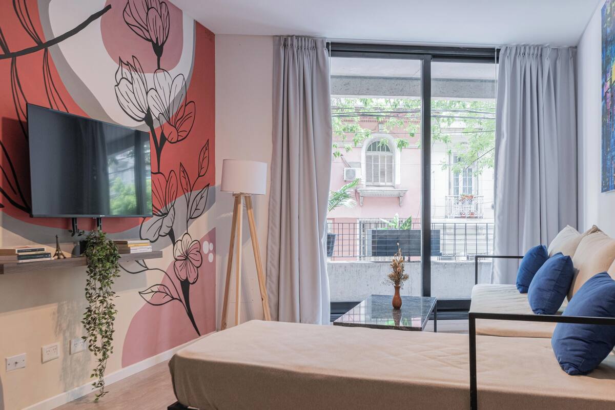 Quarto de hotel moderno com uma janela grande, televisão de tela plana, cama com almofadas azuis e um mural na parede.