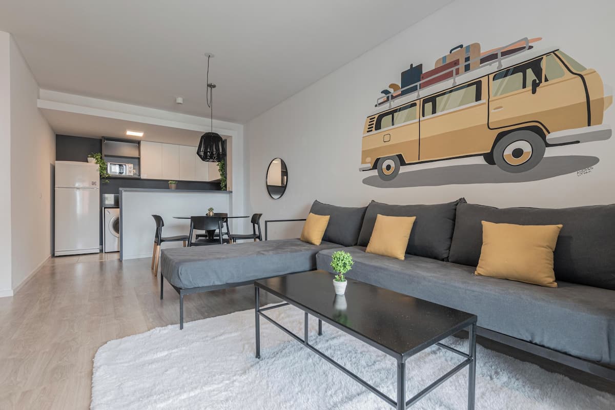 Uma sala de estar moderna com um sofá cinza, uma mesa de centro e um tapete branco. É visível, ao fundo, uma cozinha com eletrodomésticos brancos e uma área de jantar com mesa e cadeiras.