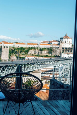 Vista de uma varanda com guarda-corpo de metal, com vista para um edifício histórico com telhados de telha vermelha e uma ponte.