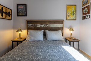 Quarto com cabeceira de madeira, duas mesas de cabeceira com luminárias, colcha estampada e quadros nas paredes.