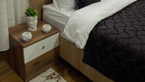 Cama bem arrumada com almofadas brancas e escuras, mesa de cabeceira de madeira com uma planta e dois objetos decorativos, e cortinas finas cobrindo uma janela.