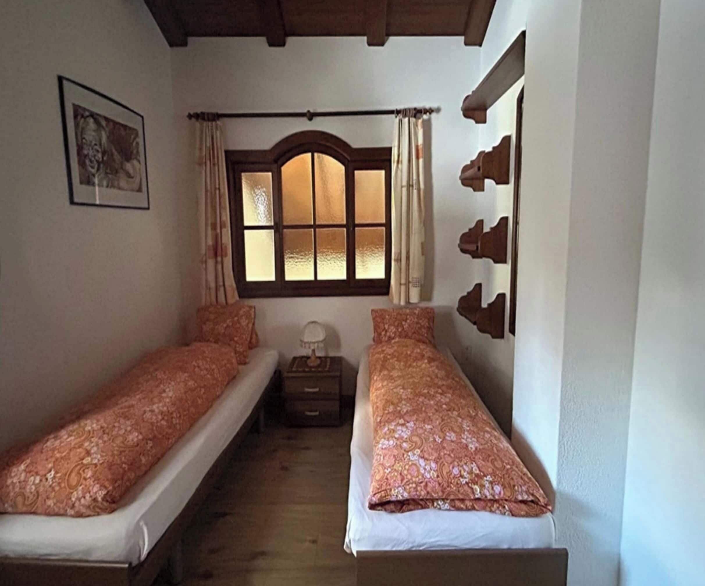 Quarto pequeno e acolhedor com duas camas de solteiro, piso de madeira, janela com cortinas e um nicho na parede.