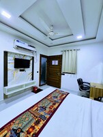 Quarto de hotel com cama, televisão na parede, ventoinha no teto, ar condicionado, porta de madeira, janela com cortinas e escrivaninha com cadeira.