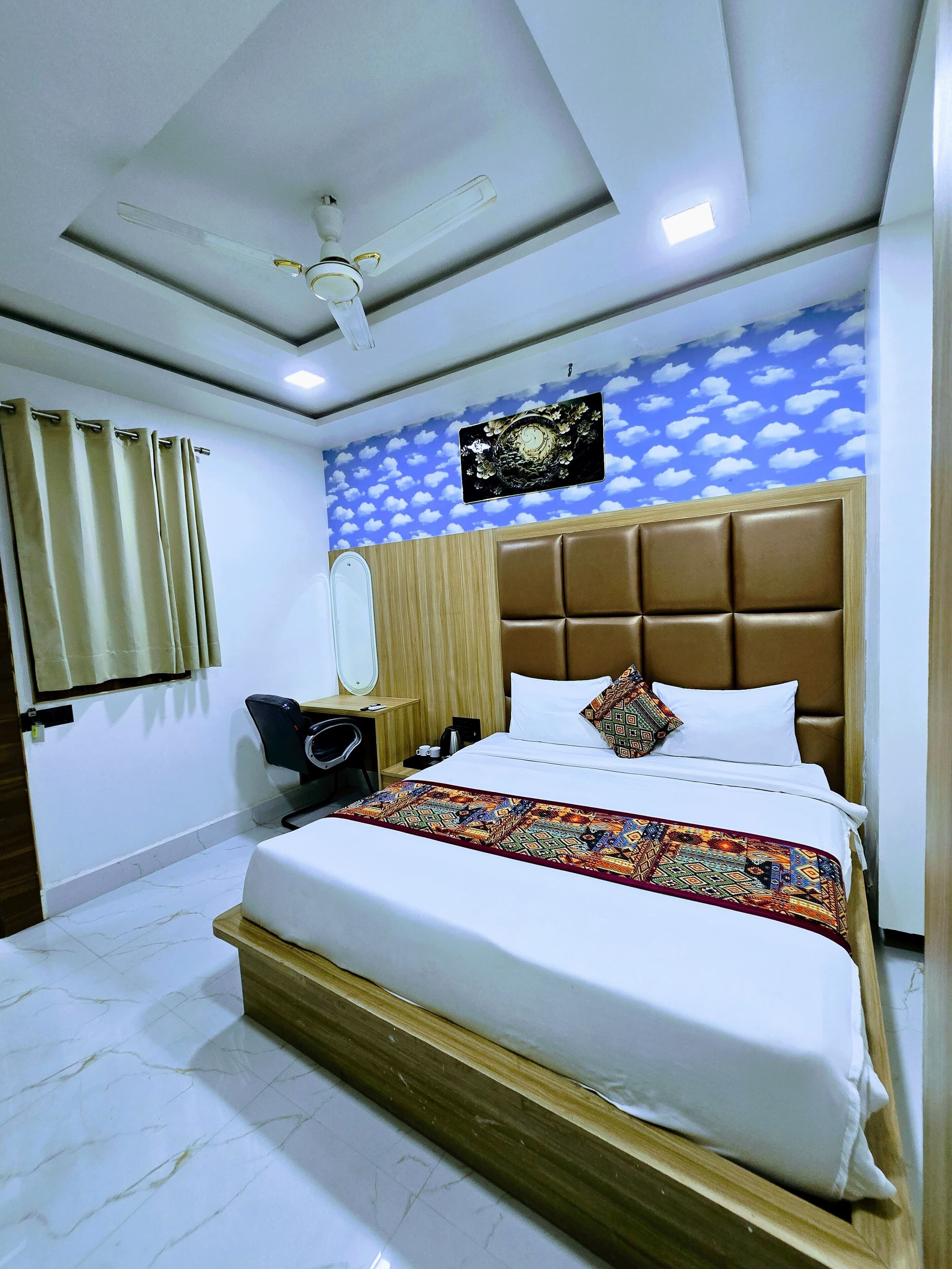 Quarto de hotel com cama, escrivaninha, cadeira, ventilador de teto e parede com padrão de nuvens.