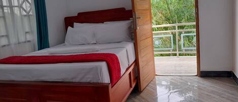 Uma cama de madeira com roupa de cama branca e um cobertor vermelho em um quarto com piso de madeira e vista para o verde através de uma porta aberta.