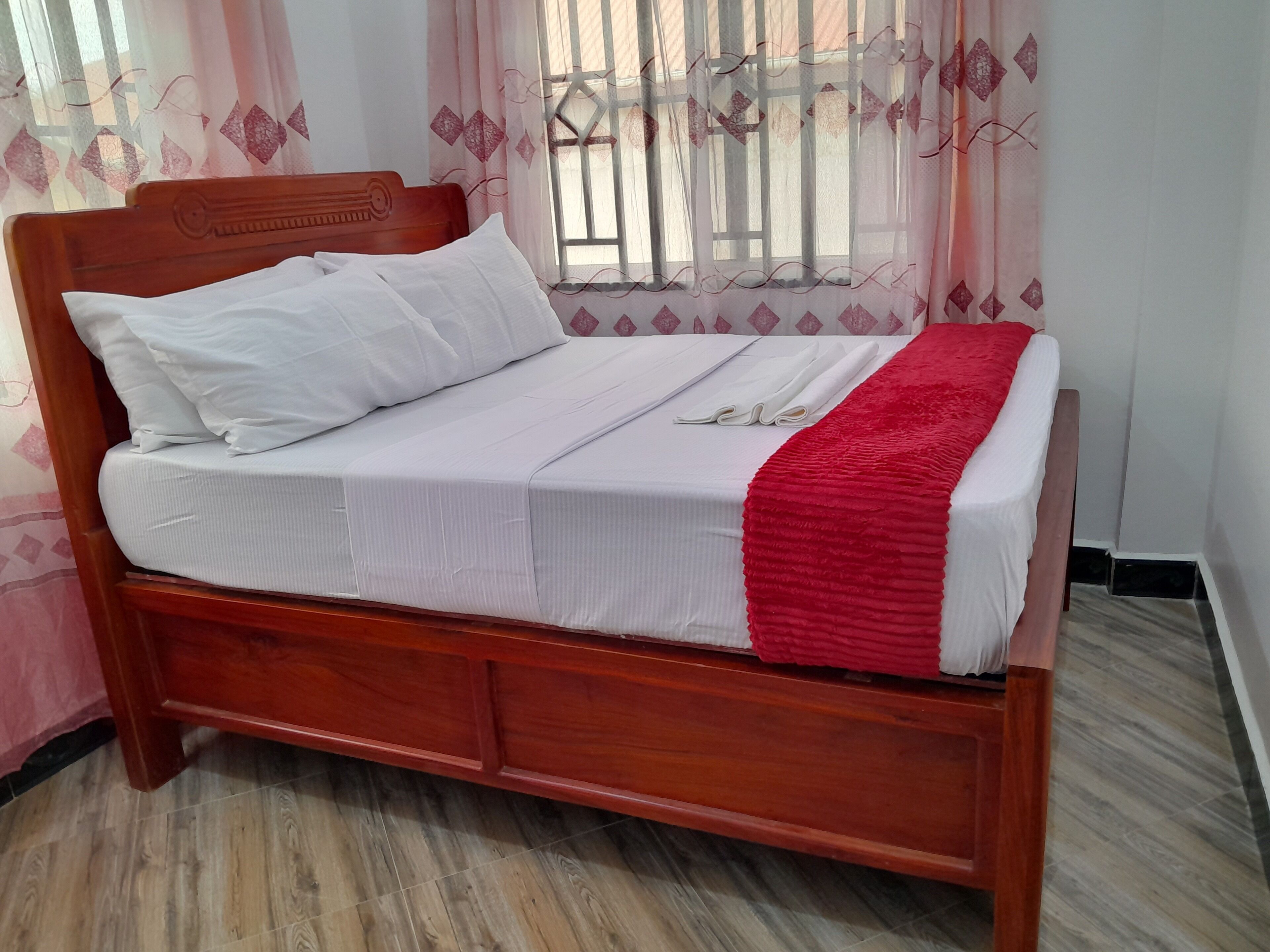 Uma cama de madeira com roupa de cama branca e um cobertor vermelho, uma janela com cortinas estampadas e um chão de azulejos.