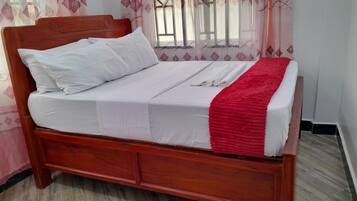 Uma cama de madeira com roupa de cama branca e um cobertor vermelho, uma janela com cortinas estampadas e um chão de azulejos.