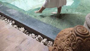 Uma mulher vestindo um roupão branco caminhando perto de uma fonte com vários níveis em um amplo pátio interno com vigas de madeira e piso de pedra.