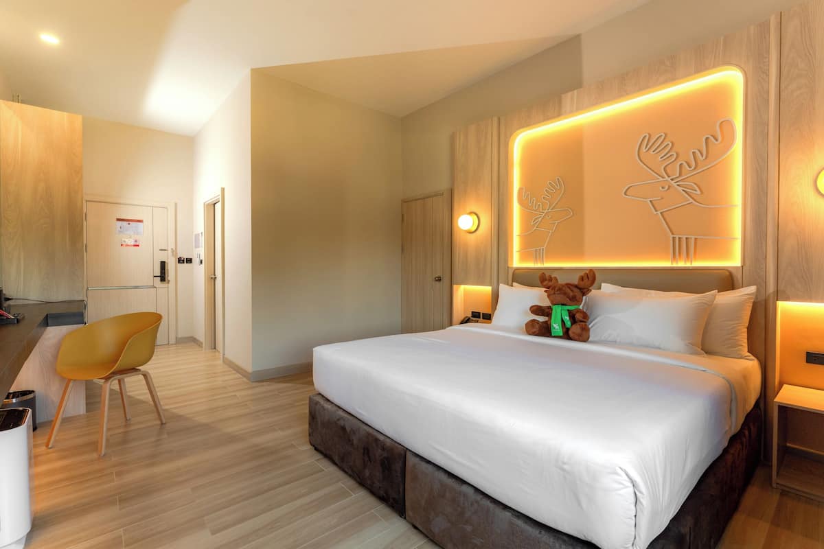 Quarto de hotel moderno com uma cama grande, cabeceira de madeira com design de alce neon, um pequeno espaço de estar com uma cadeira amarela e uma cozinha compacta ao fundo.