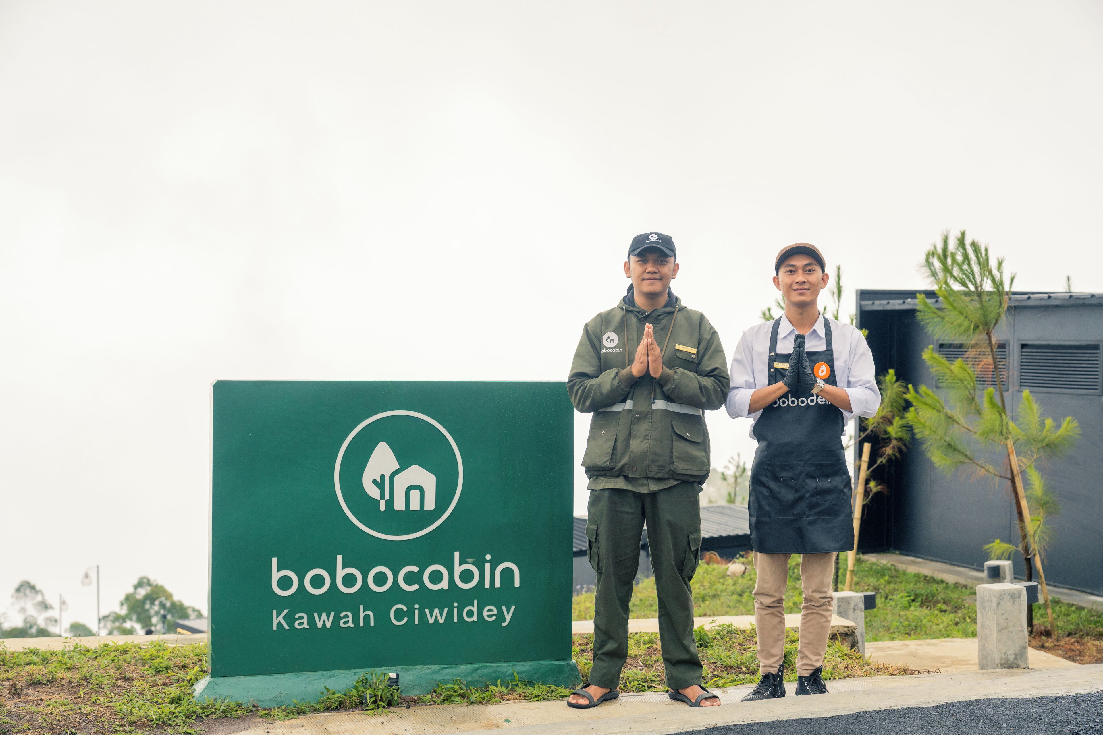 Duas pessoas em frente a uma placa de Bobocabin Kawah Ciwidey.