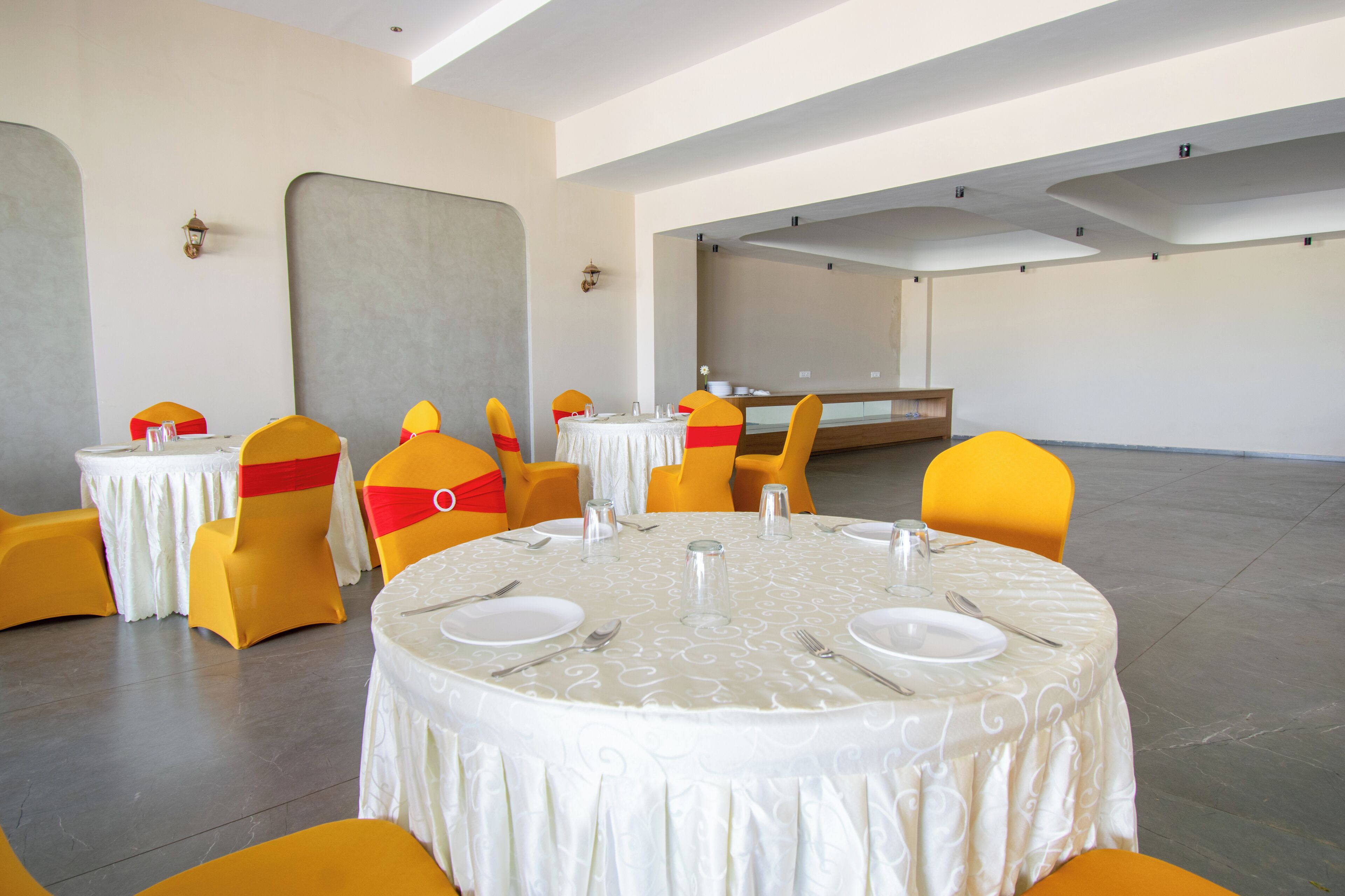Uma área de refeições com mesas redondas, cadeiras amarelas e toalhas de mesa brancas.