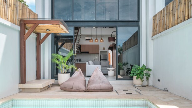 Uma área de piscina moderna ao ar livre com pérgola de madeira, almofadas para sentar e plantas em vasos.