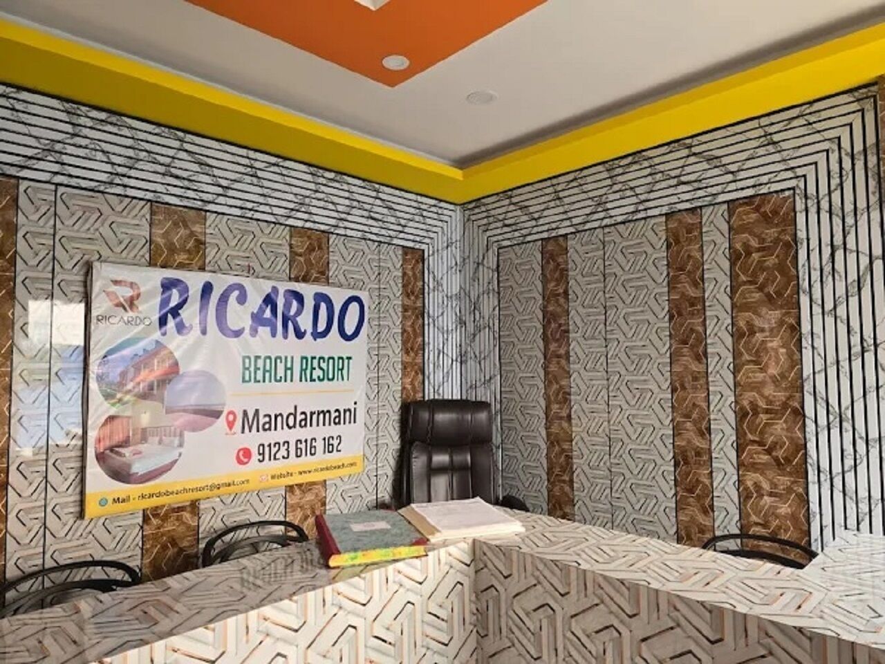 Uma sala de conferências com um banner do Ricardo Beach Resort, uma cadeira de couro preto e papel de parede estampado.