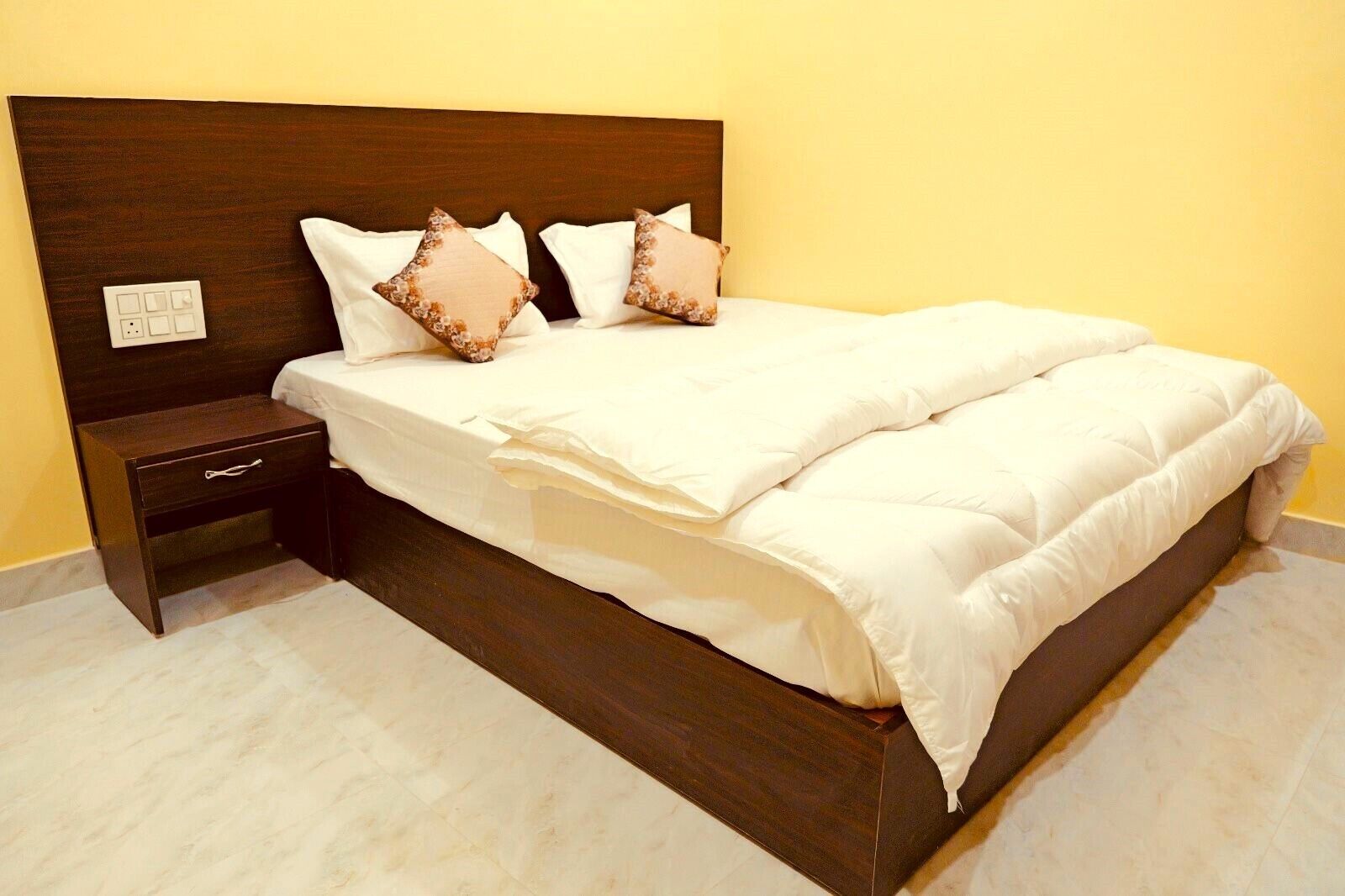 Uma cama bem arrumada com roupa de cama branca e almofadas decorativas, uma cabeceira de madeira e uma pequena mesa de cabeceira com uma gaveta.