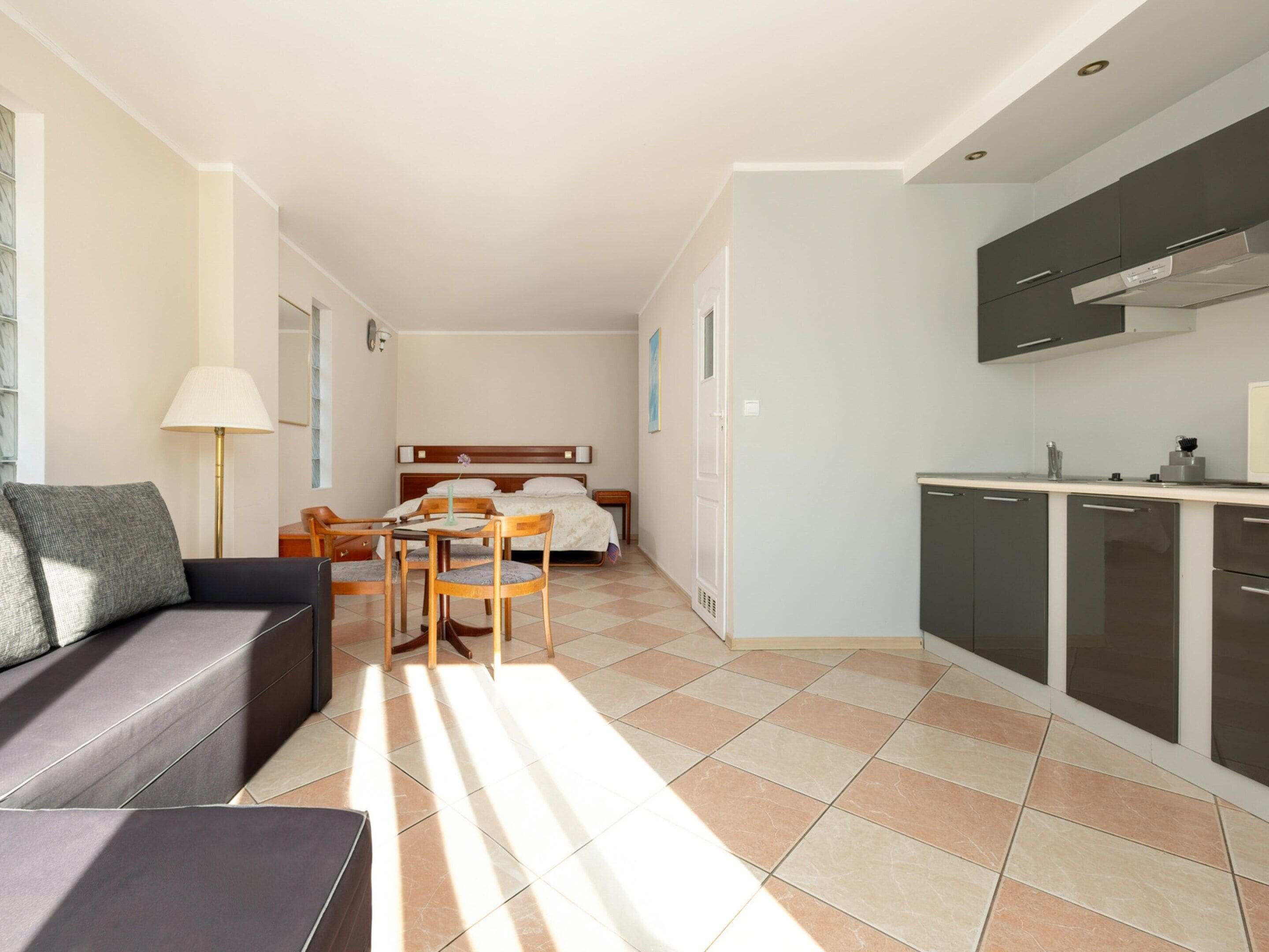 Quarto de hotel moderno com cama, sofá, mesa de jantar com cadeiras e cozinha compacta.