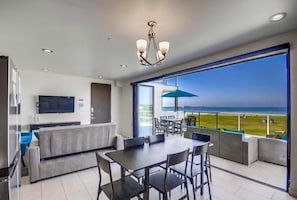 Uma cozinha moderna com uma TV de tela plana, uma área de jantar com mesa e cadeiras, e uma grande janela com vista para o oceano.
