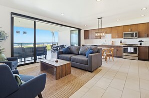 Cozinha moderna com eletrodomésticos de aço inoxidável, área de jantar com mesa e cadeiras de madeira, sofá cinza com almofadas azuis e uma grande porta de vidro deslizante que dá para uma varanda com vista para a praia.