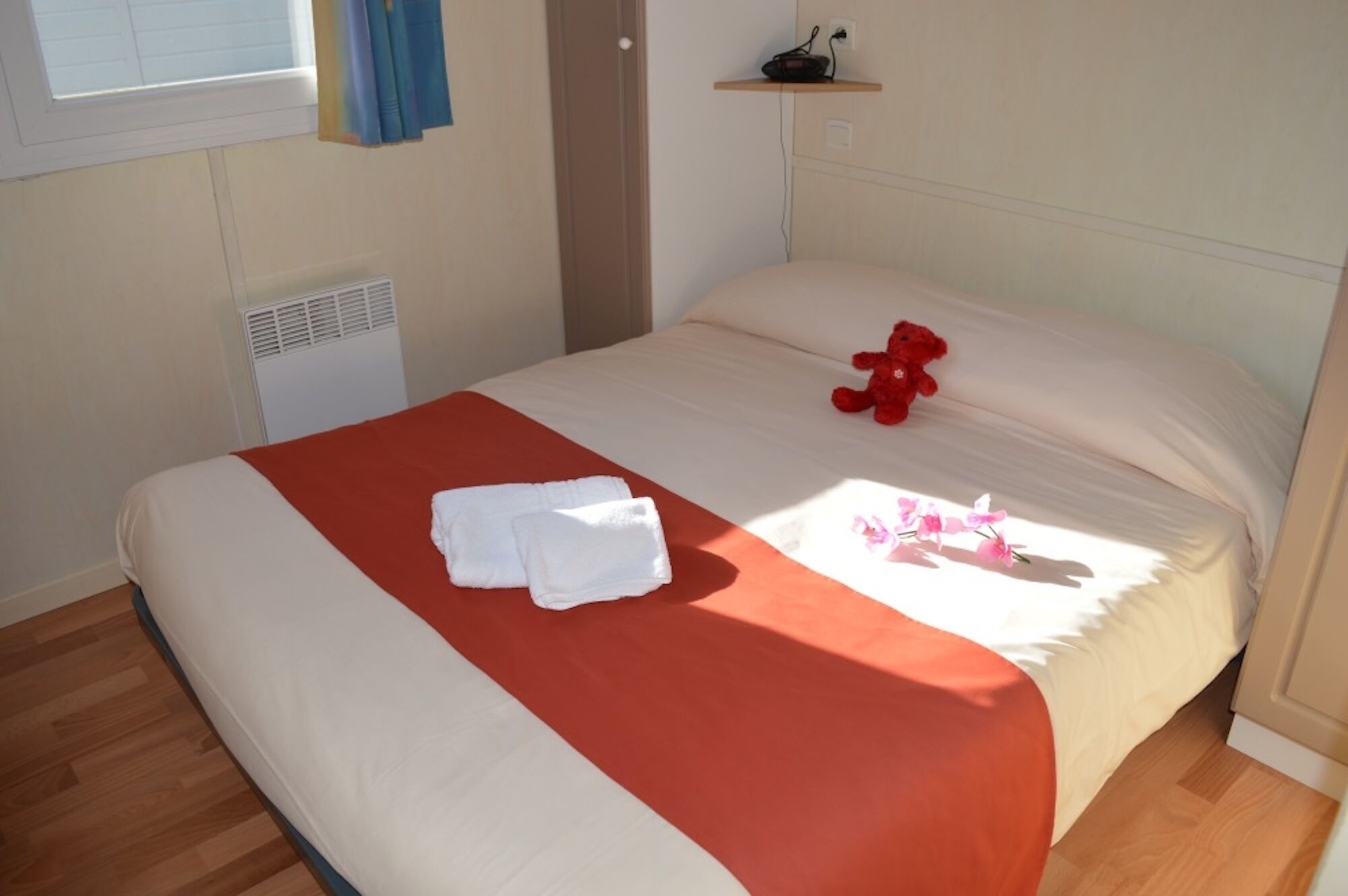 Quarto de hotel com uma cama, um cobertor vermelho, uma toalha branca, um ursinho de pelúcia vermelho e uma flor rosa.