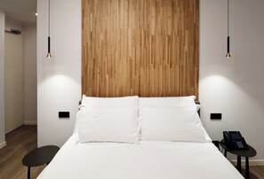 Quarto de hotel com cabeceira de madeira, roupa de cama branca, mesa de cabeceira preta com um telefone e iluminação pendente.