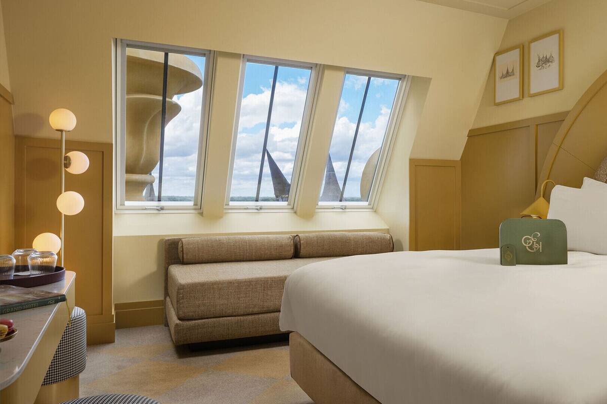 Quarto de hotel com cama, área de estar, escrivaninha com abajur e janela com vista para uma ponte.