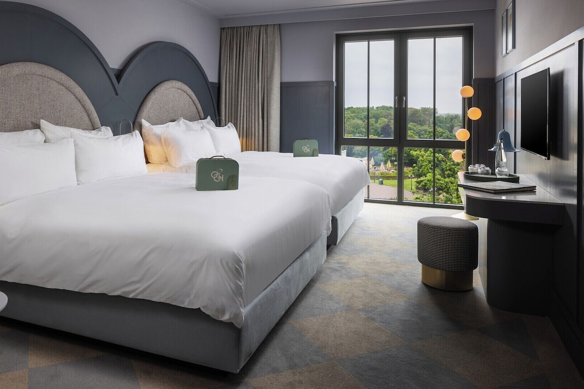 Quarto de hotel com uma cama grande, uma escrivaninha, uma cadeira, uma televisão e vista para o verde pela janela.