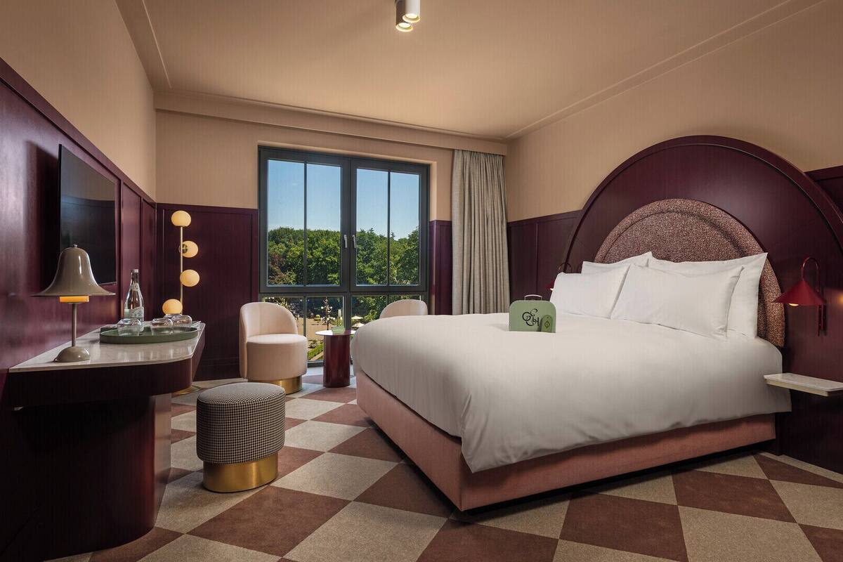 Quarto de hotel com uma cama grande, uma escrivaninha com abajur, uma cadeira e vista para o verde pela janela.