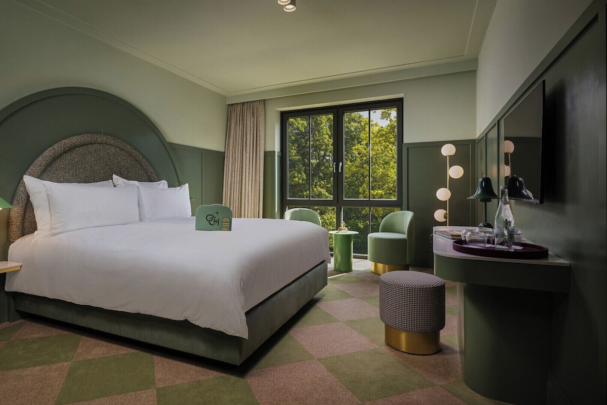 Quarto de hotel moderno com uma cama grande, uma toucador com espelhos, uma janela com vista para árvores e uma parede de destaque verde.