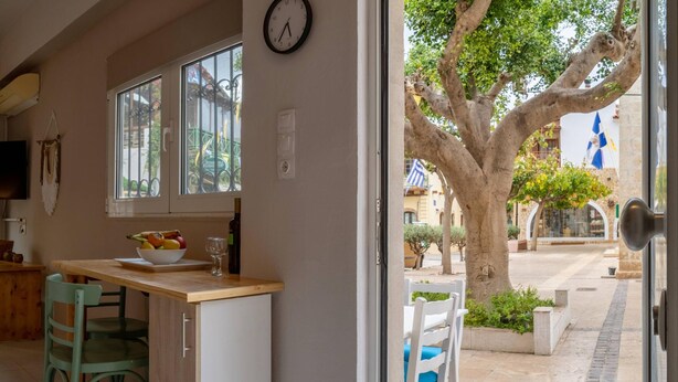 Quarto com uma mesa de madeira, uma tigela de frutas e uma cadeira. Há uma janela com grades e vista para o exterior.