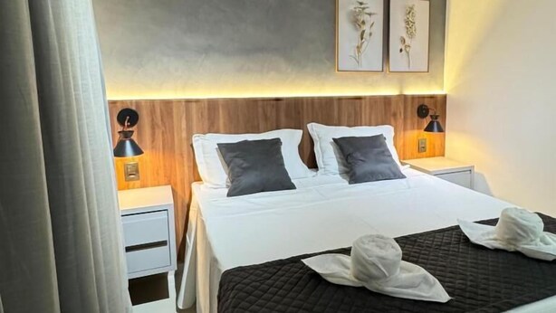 Quarto de hotel com uma cama grande, duas mesas de cabeceira com abajures, uma cabeceira de madeira e duas obras de arte penduradas na parede.