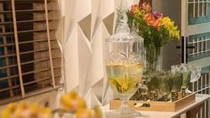 Uma área de refeições com um assento estofado de couro marrom, uma mesa redonda com um vaso de flores e uma janela com persianas.