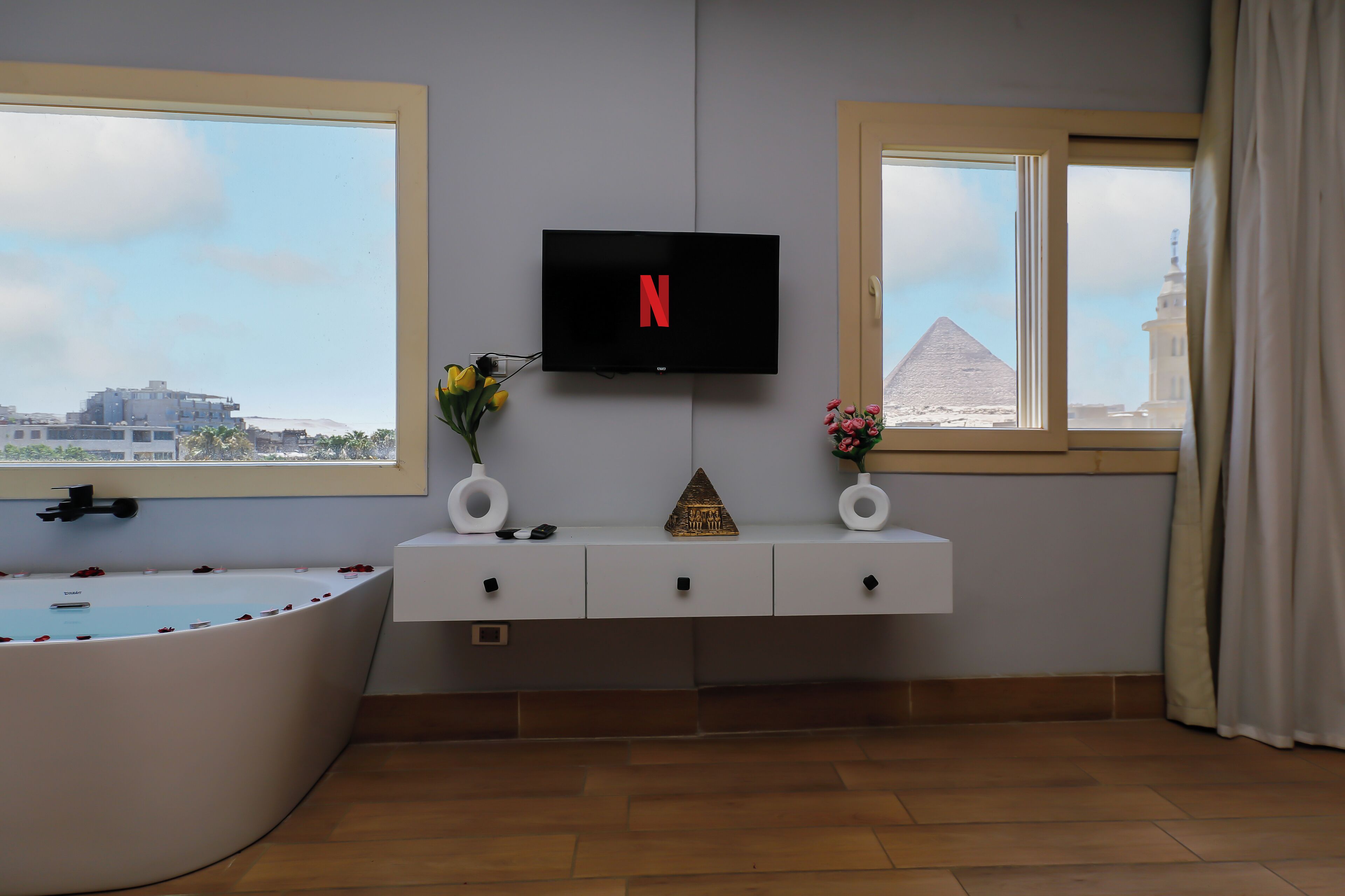 Uma casa de banho moderna com uma banheira grande, uma TV Netflix na parede e vista para uma pirâmide através de uma janela.