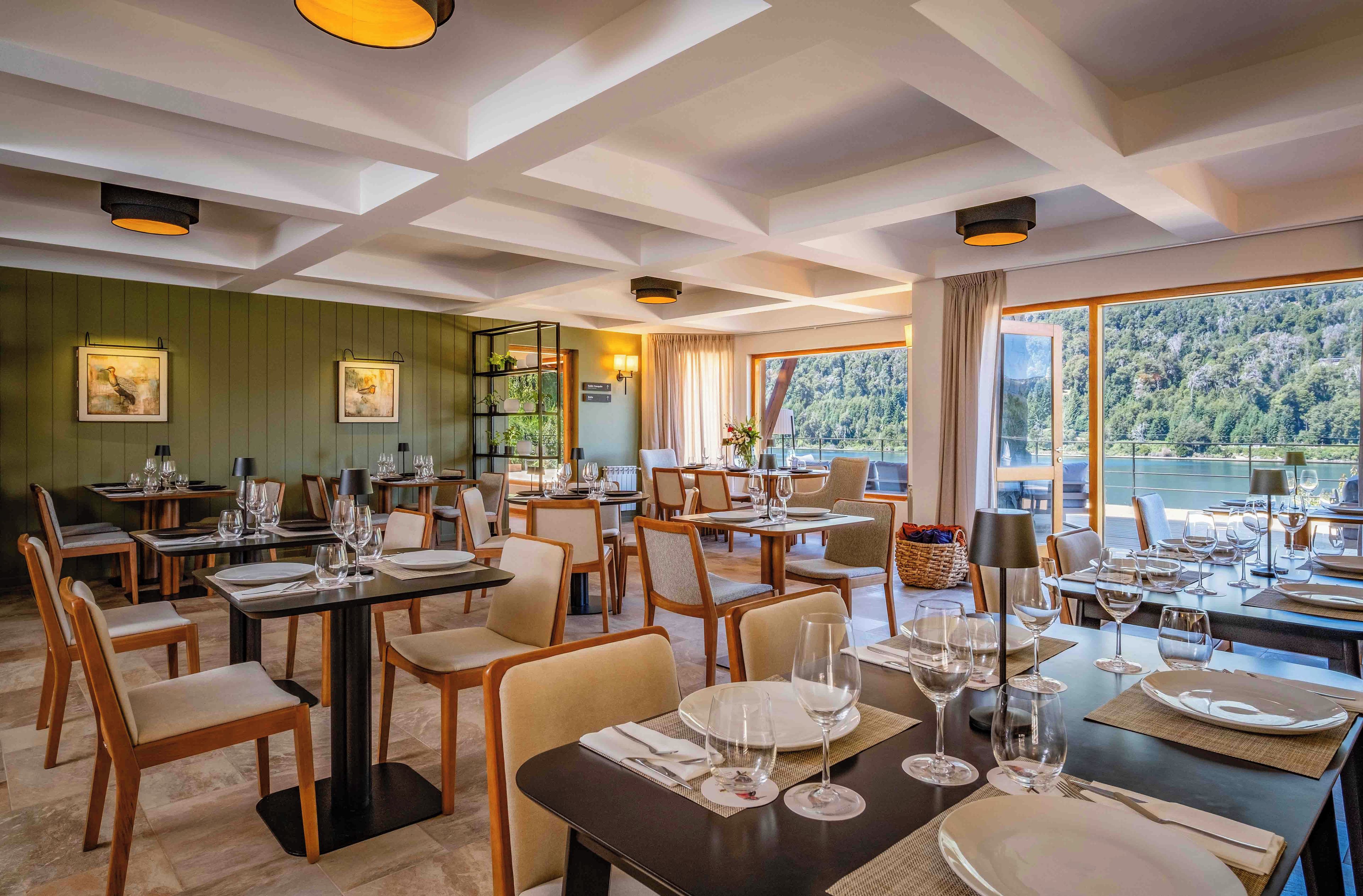 Restaurante com várias mesas, cadeiras e vista para uma área arborizada através de grandes janelas.