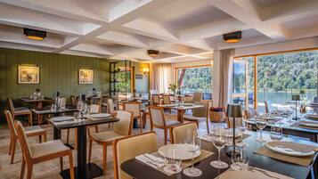 Restaurante com várias mesas, cadeiras e vista para uma área arborizada através de grandes janelas.