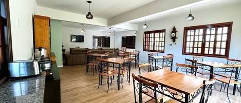 Uma sala de jantar ampla com mesas e cadeiras de madeira, área de cozinha com fogão e geladeira, e grandes janelas que proporcionam luz natural.