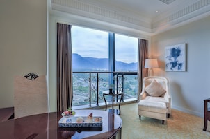 Quarto de hotel com uma janela grande, uma cadeira, uma mesa com bandeja e um abajur.