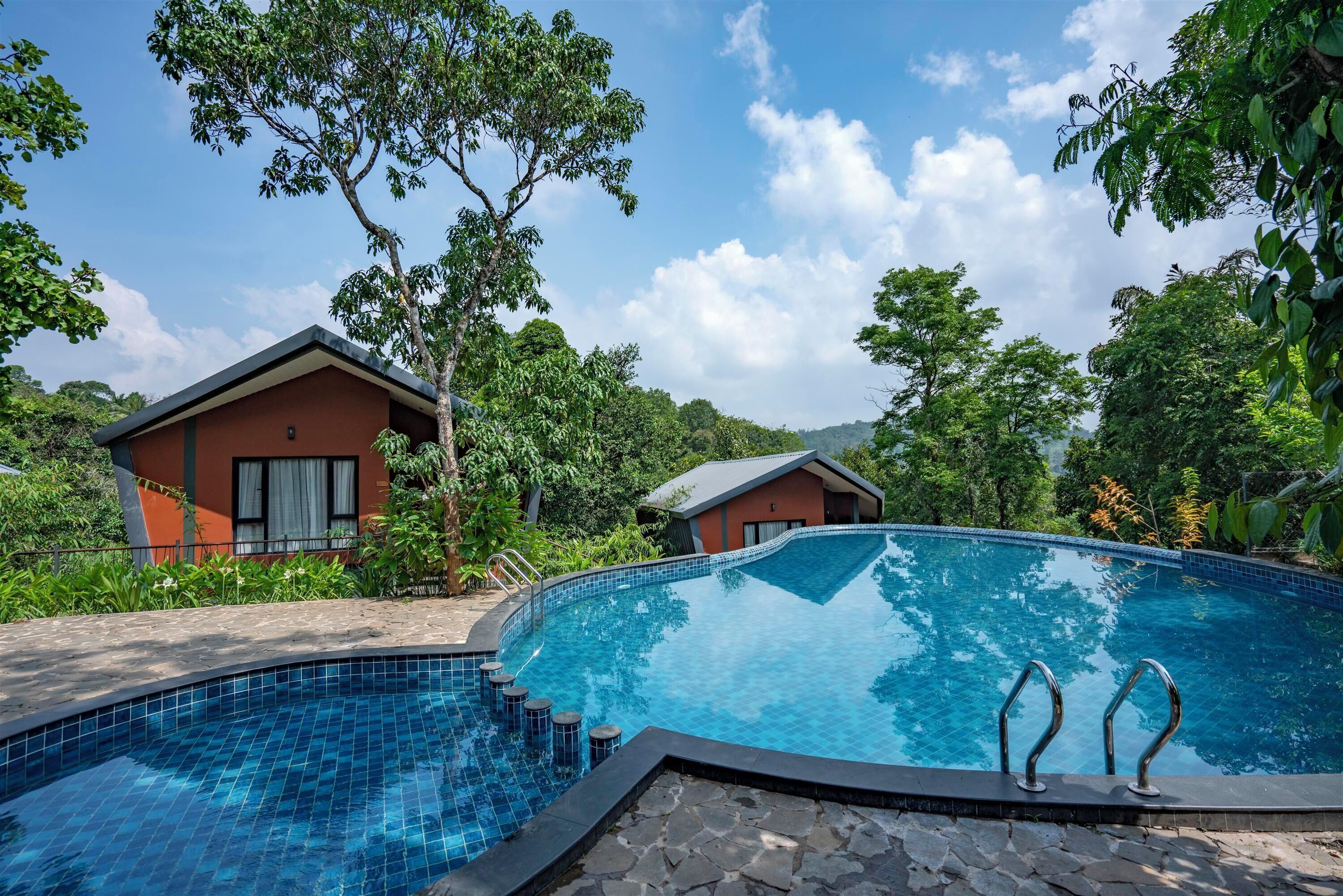 Piscina com borda de pedra, rodeada por vegetação exuberante e dois edifícios pequenos.