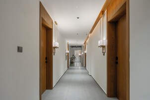 Um corredor comprido e estreito, com portas de madeira de ambos os lados, iluminação embutida e chão de azulejos.