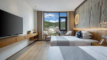 Quarto de hotel moderno com uma grande TV de tela plana, uma mesa consola de madeira, duas camas, uma área de estar com uma poltrona e vista para a cidade através de grandes janelas.
