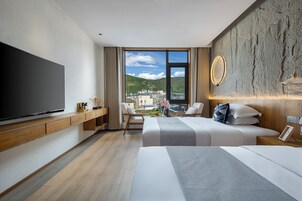 Quarto de hotel moderno com uma grande TV de tela plana, uma mesa consola de madeira, duas camas, uma área de estar com uma poltrona e vista para a cidade através de grandes janelas.