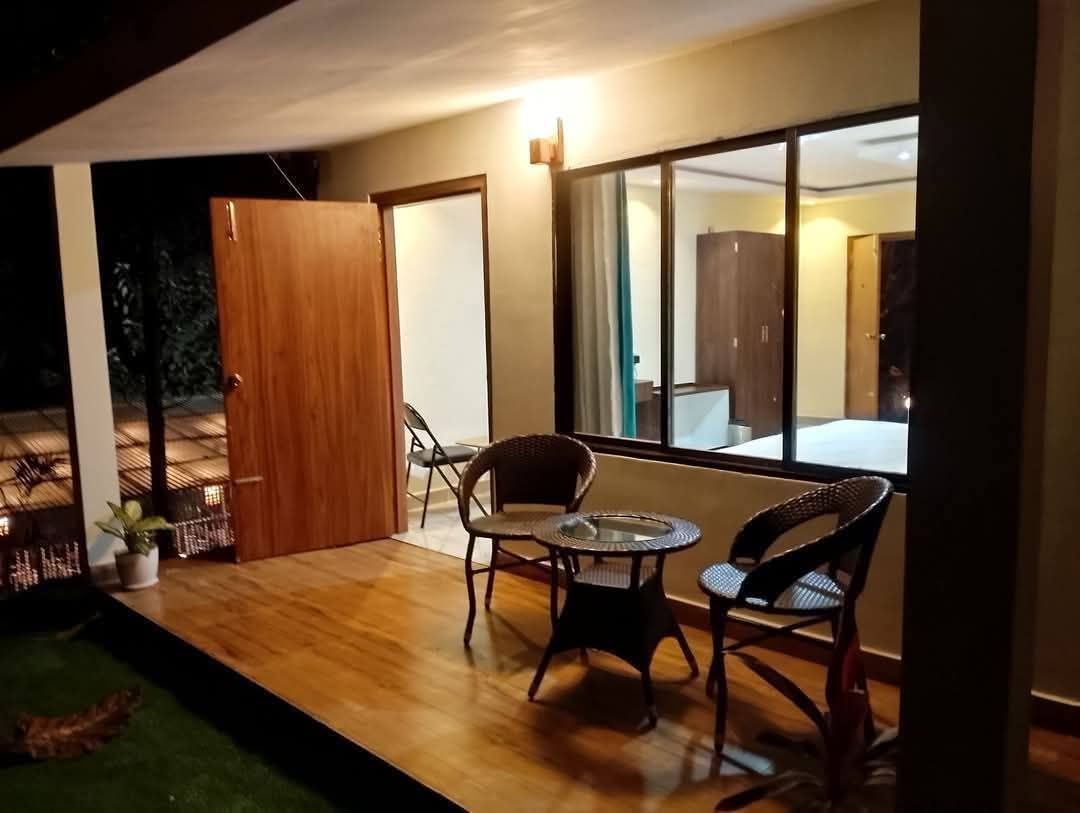 Quarto com piso de madeira, uma mesa redonda pequena, duas cadeiras de vime e uma janela grande.